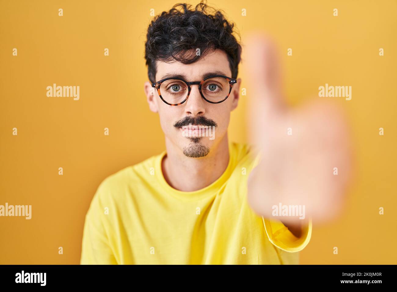 Hispanic man wearing glasses standing over yellow background smiling ...