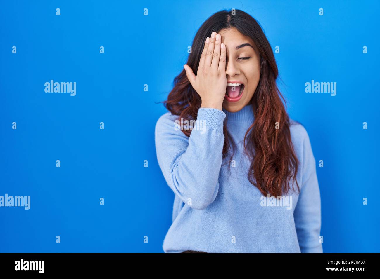 Hispanic young woman standing over blue background covering one eye ...