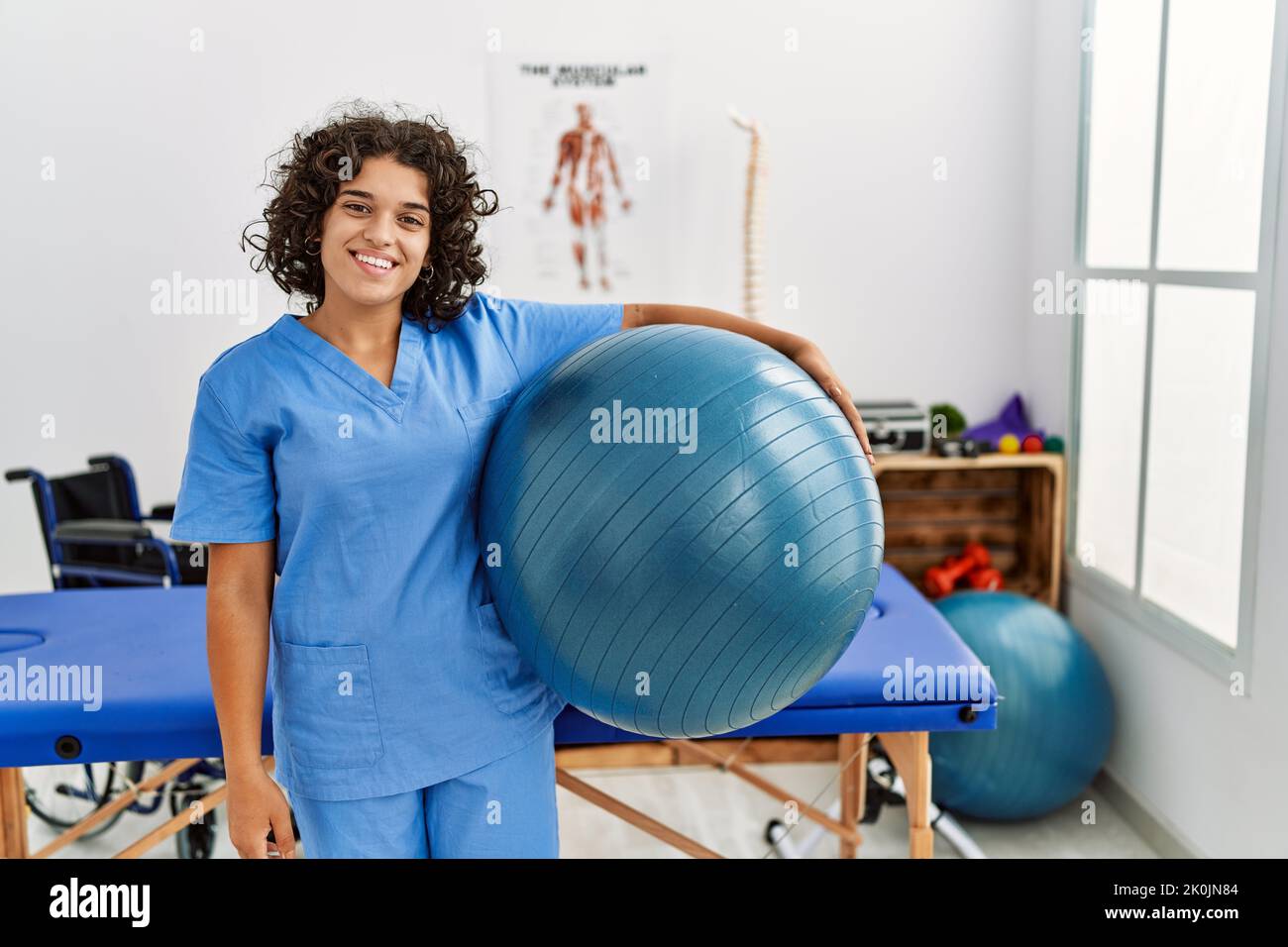 Young hispanic woman wearing physio therapist uniform holding fit ball ...