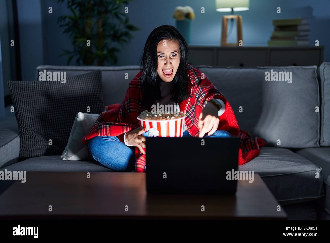 Hispanic woman eating popcorn watching a movie on the sofa pointing ...