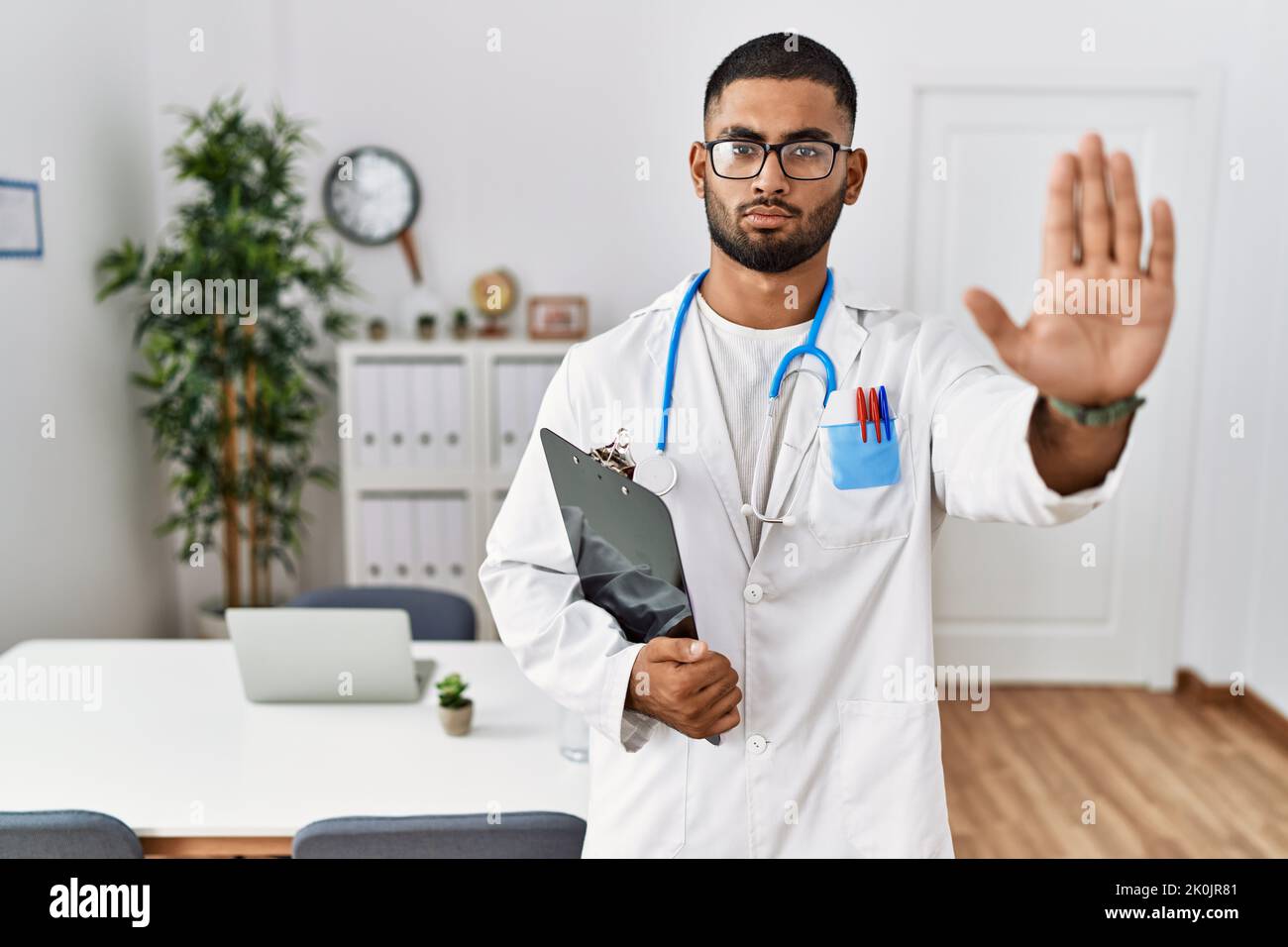 Young indian man wearing doctor uniform and stethoscope doing stop sing ...