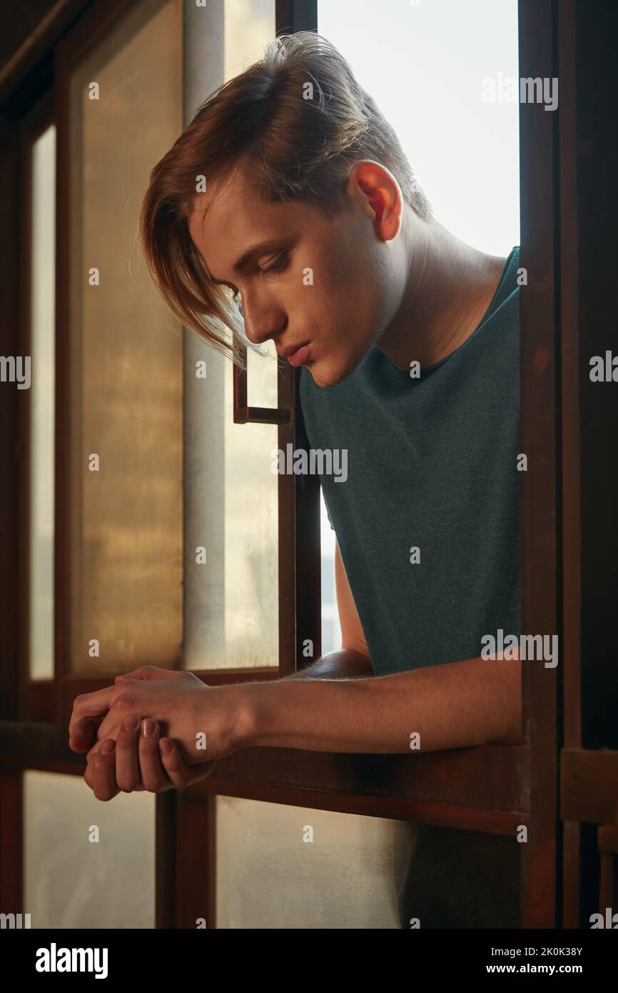 Serious young blond male with long bangs looking out open window from ...