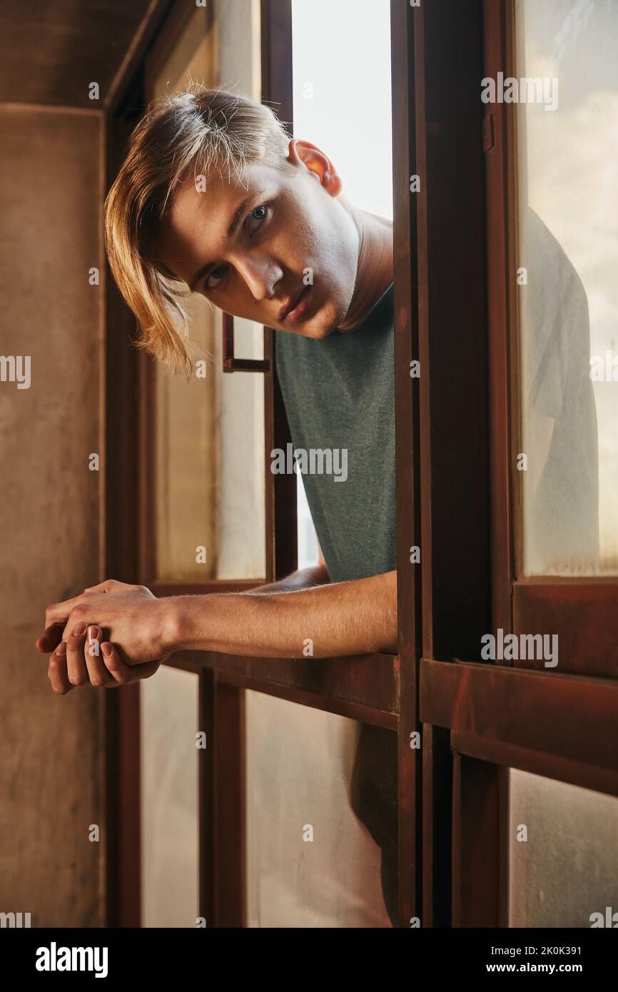 Serious young blond male with long bangs looking out open window from ...