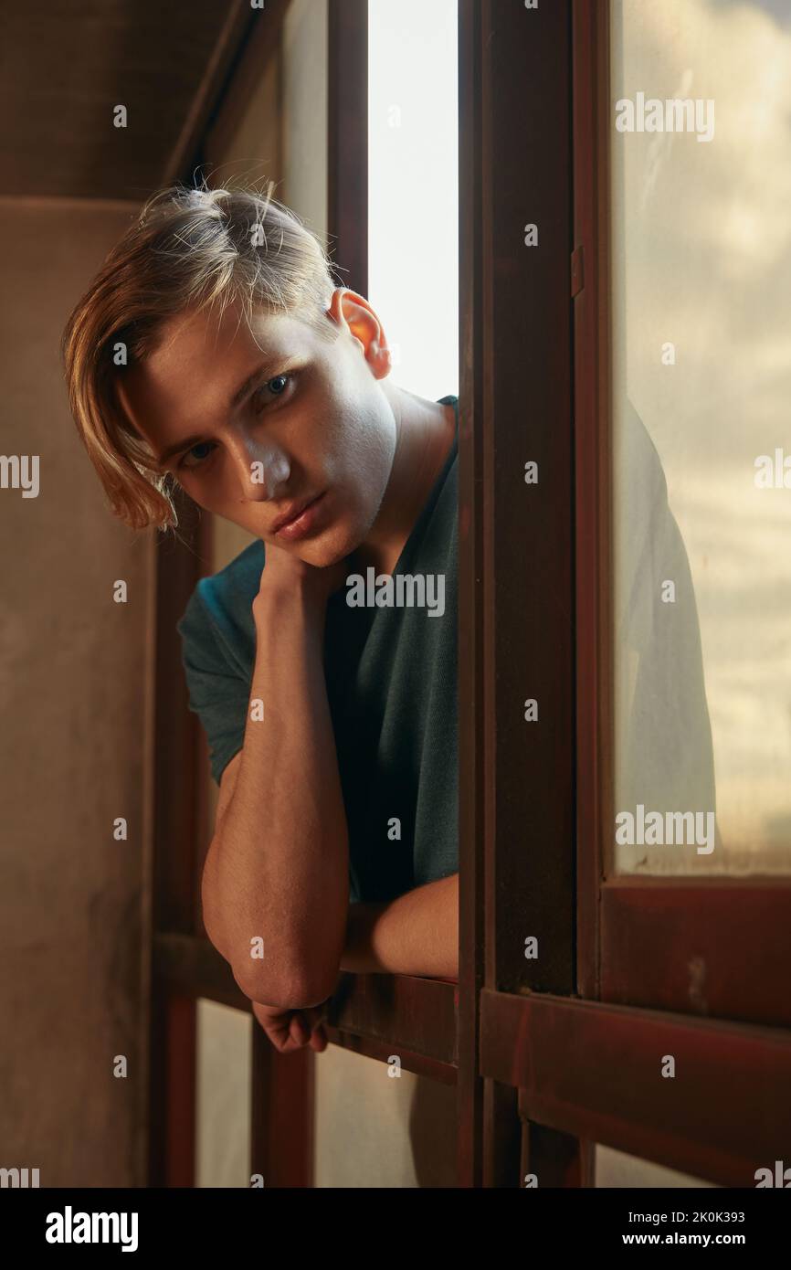 Serious young blond male with long bangs looking out open window from ...