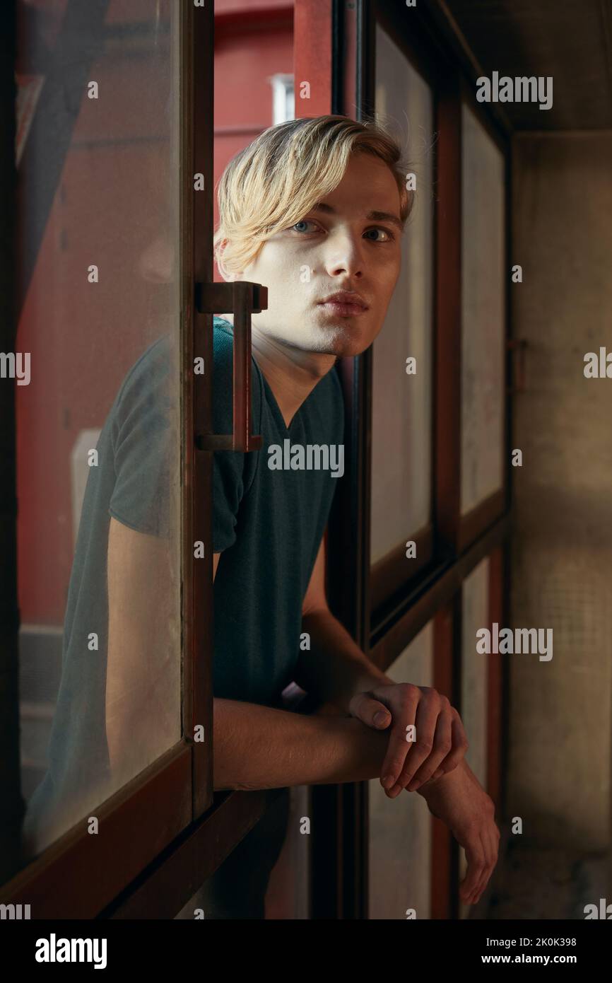 Serious young blond male with long bangs looking out open window from ...