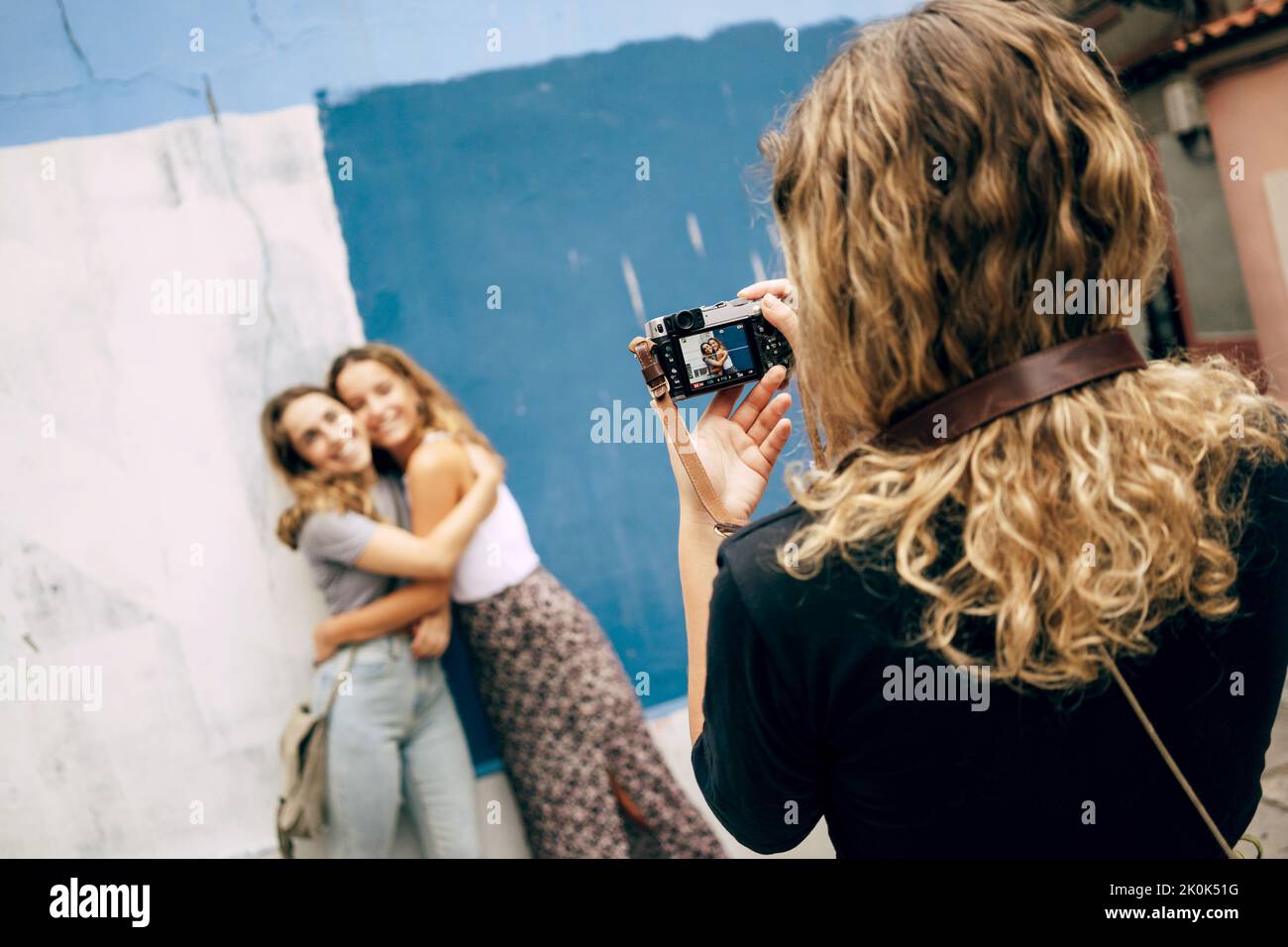 Charming friends hugging posing against wall while woman taking picture ...