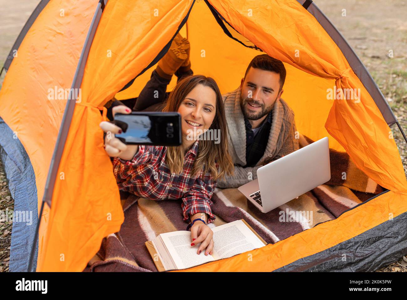 Young happy couple taking selfie on smartphone while reading and browsing on laptop during ...
