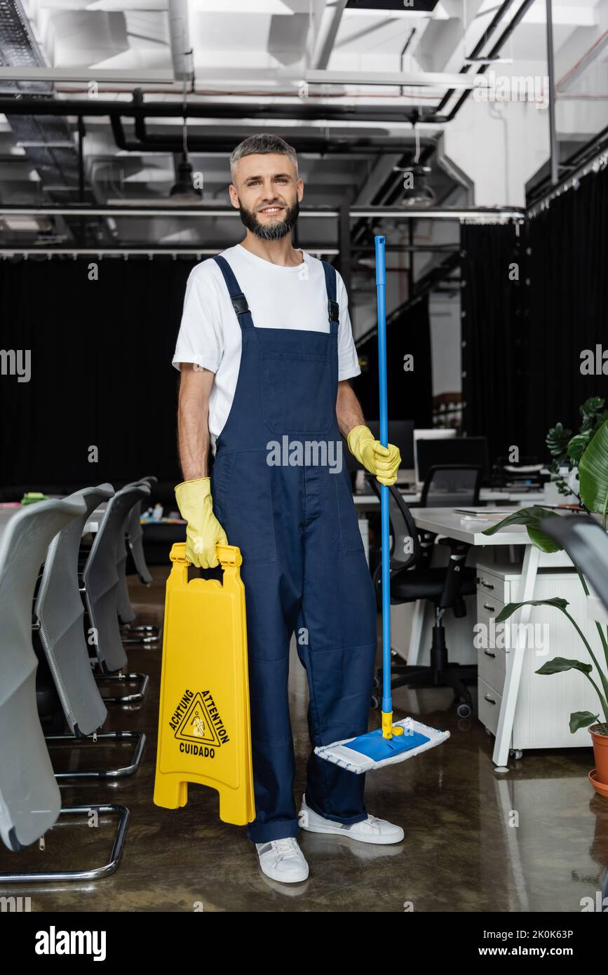 full length of man in overalls holding mop and attention sign board ...