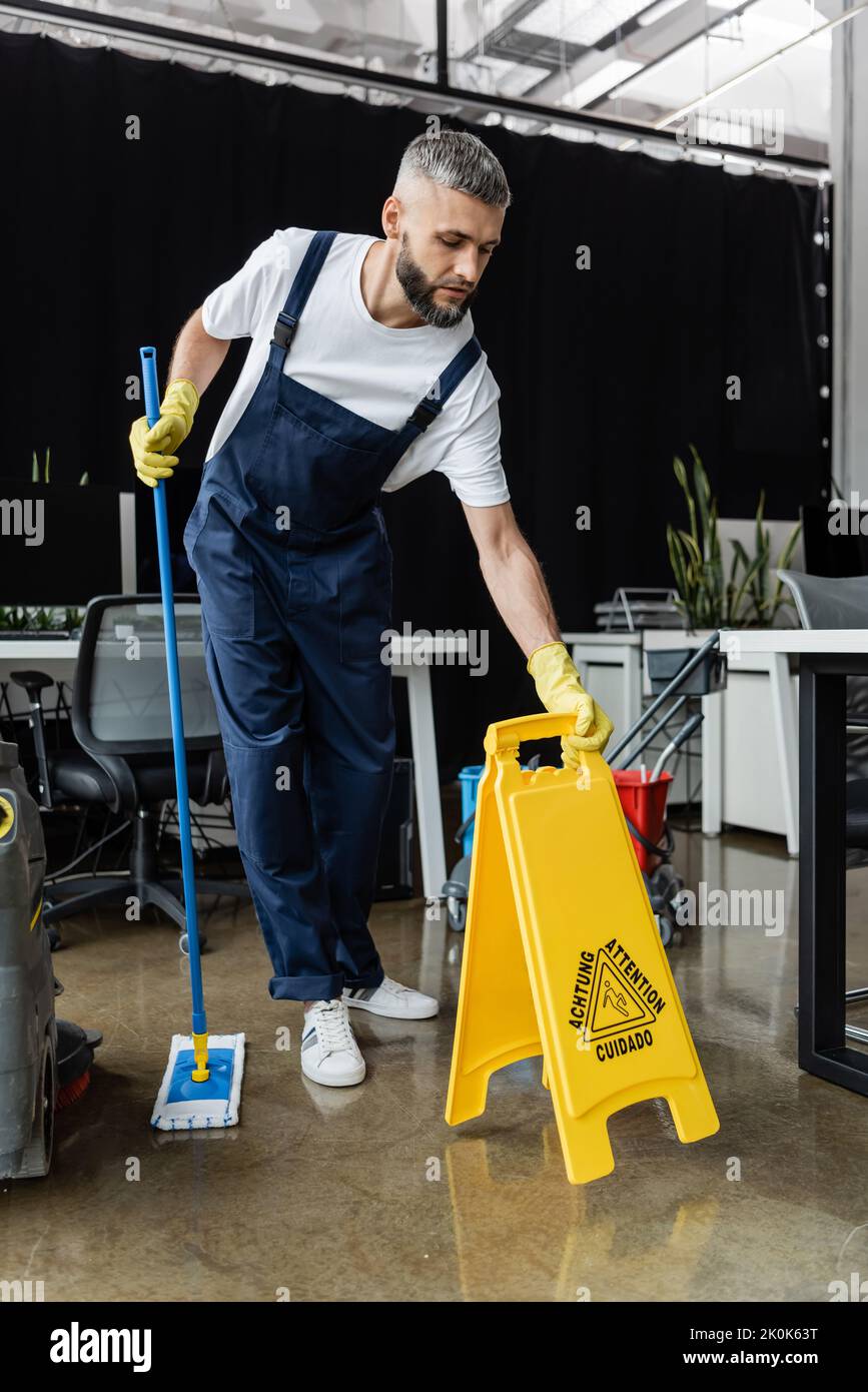 professional cleaner in overalls holding mop and caution sign board in ...
