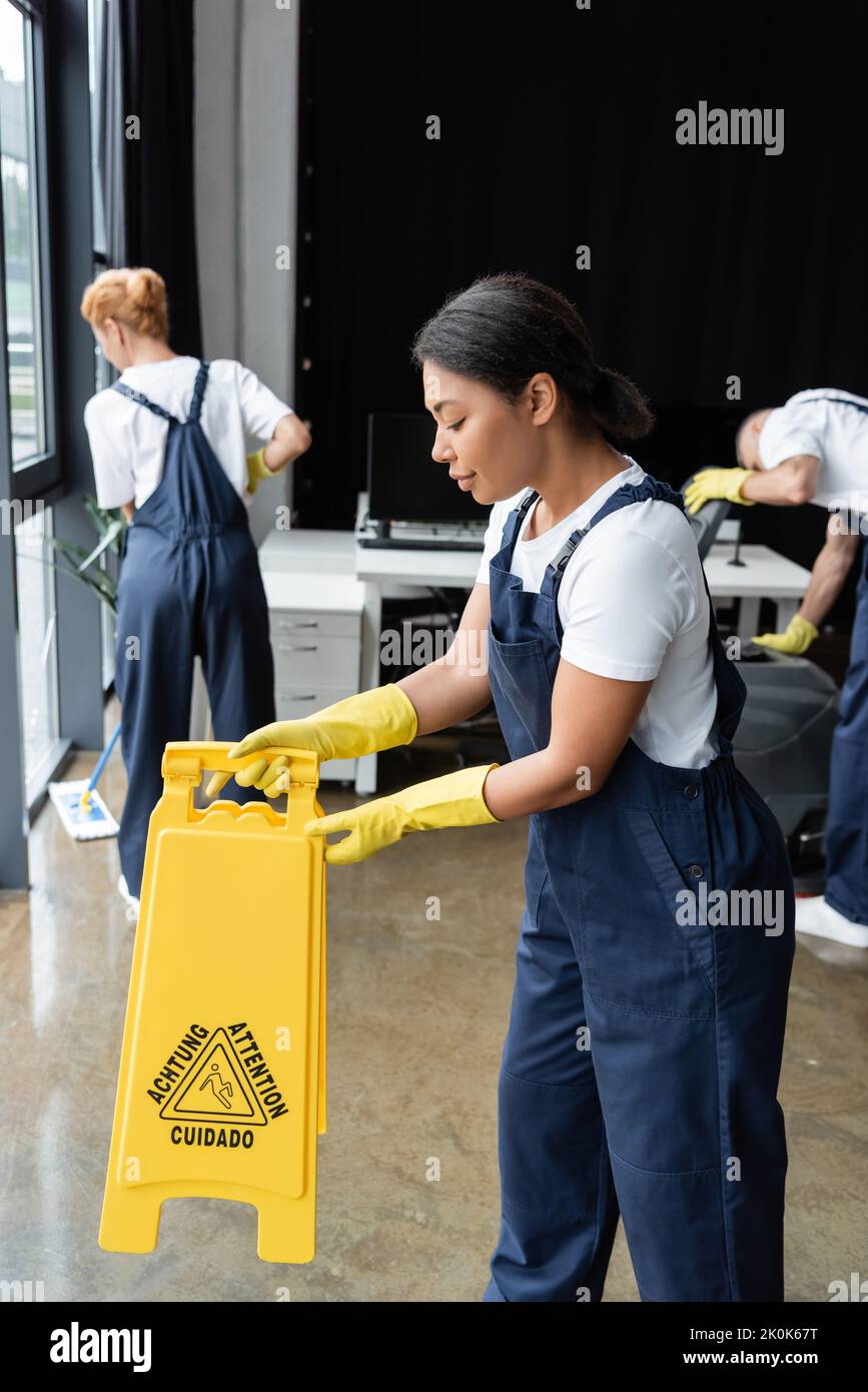 young bi-racial woman holding warning board near colleagues cleaning ...