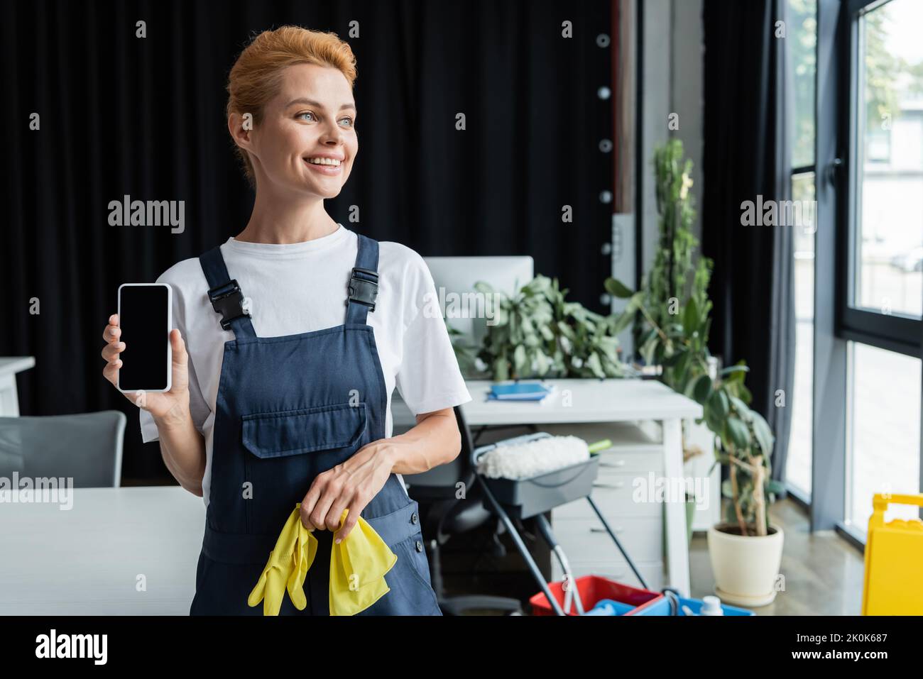 cheerful woman in overalls holding smartphone with blank screen while ...