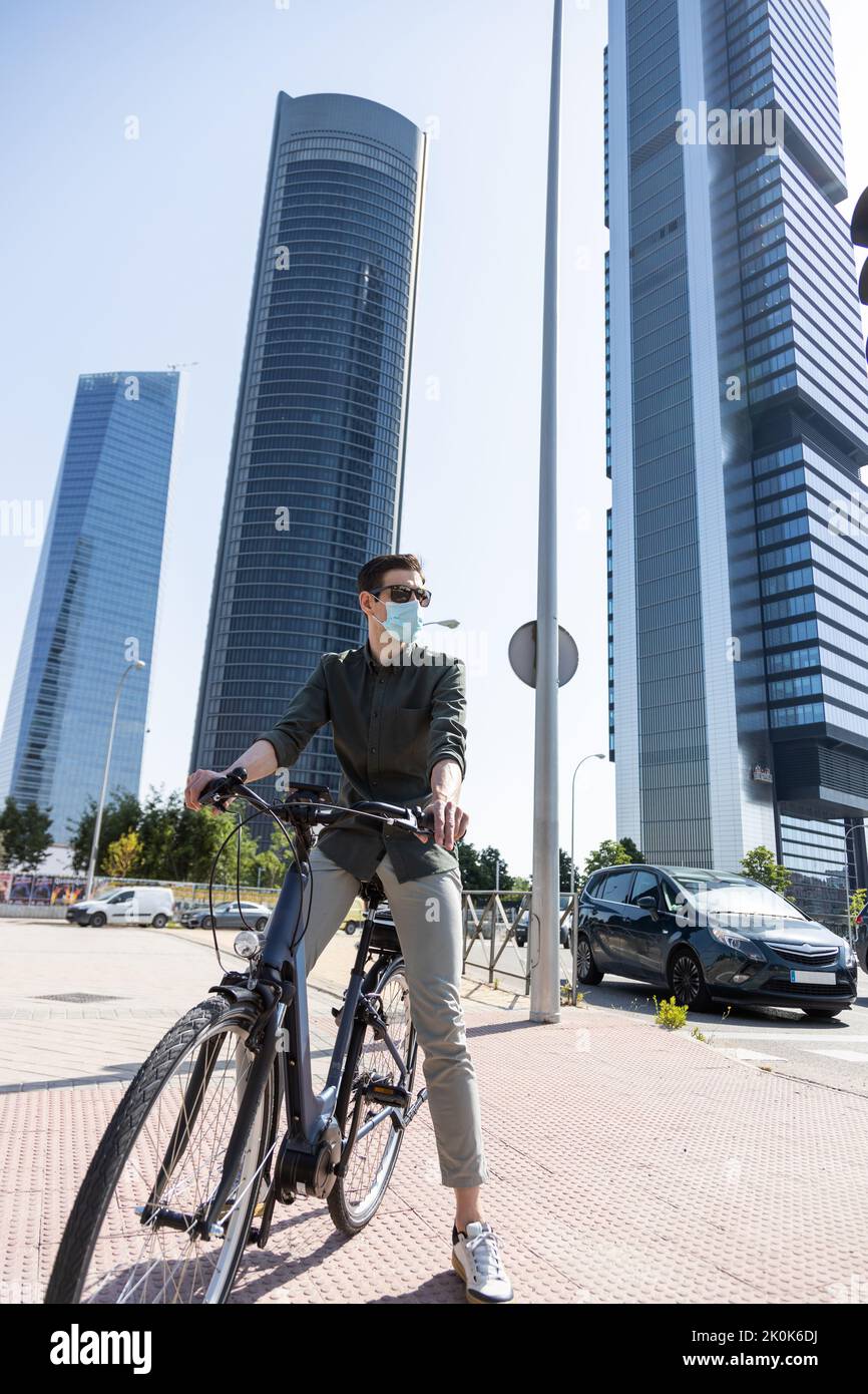 Male in protective mask riding contemporary bike along street while commuting to work in ...