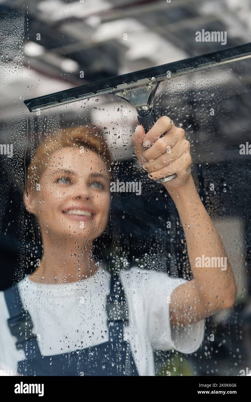 professional cleaner with window squeegee smiling near wet glass in ...