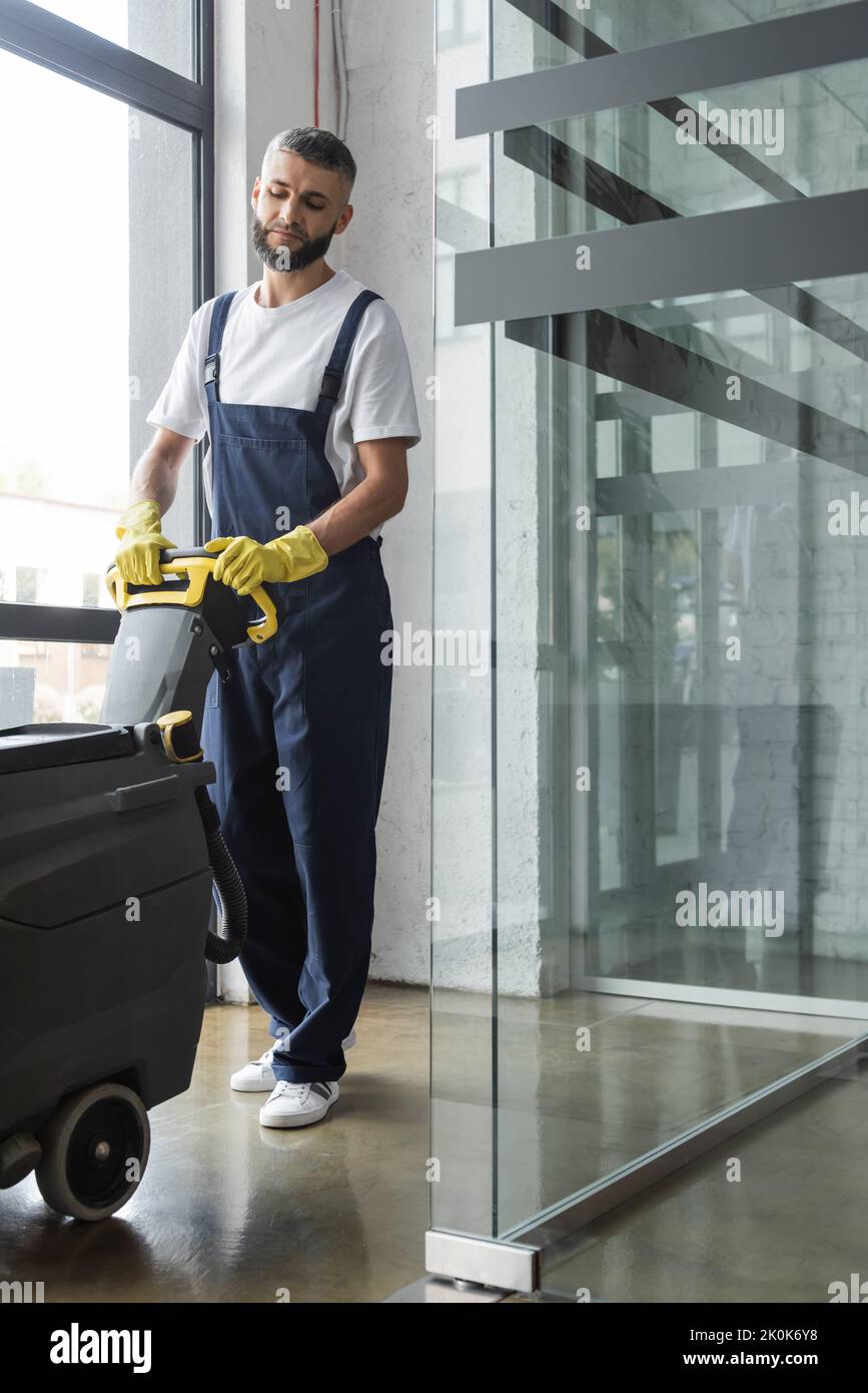 full length of man in overalls cleaning office with electrical floor ...