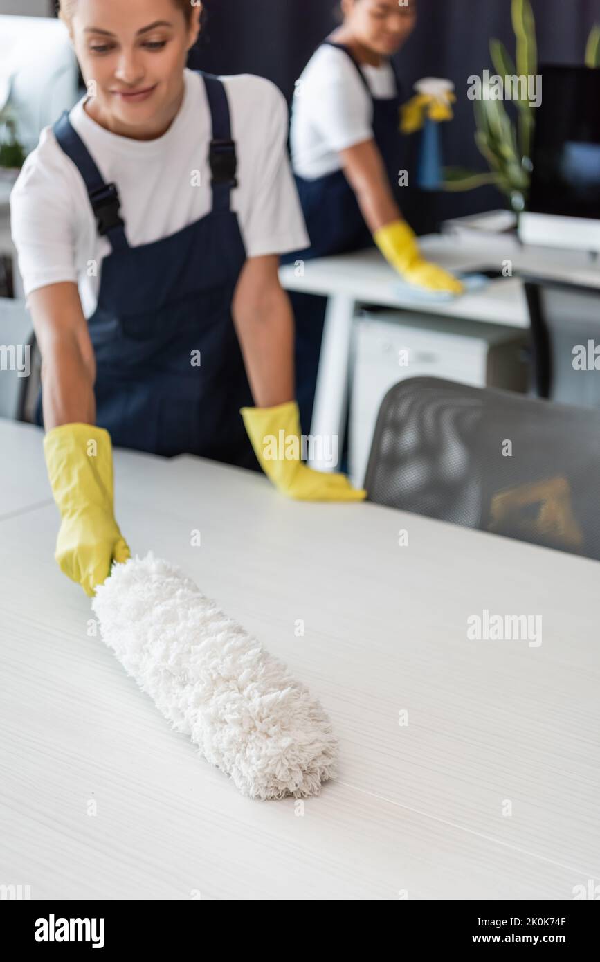 smiling woman cleaning desk with dust brush near blurred mixed race ...