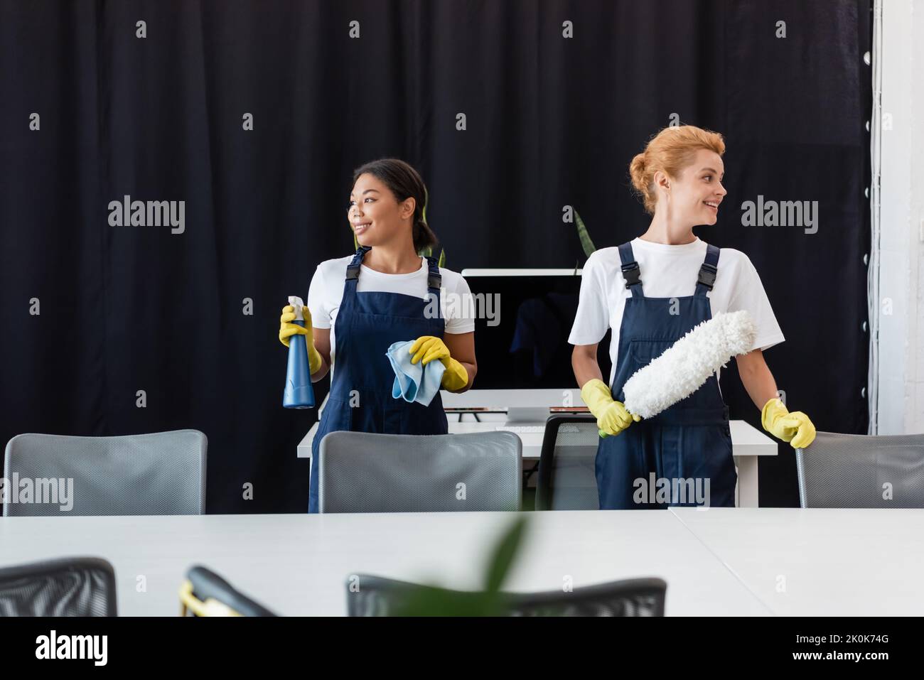 interracial women in overalls holding cleaning supplies and smiling in ...