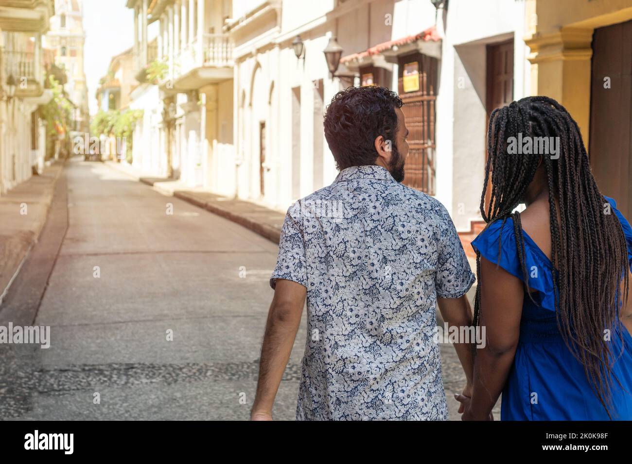 Back view of multiracial couple holding hands and walking along street ...