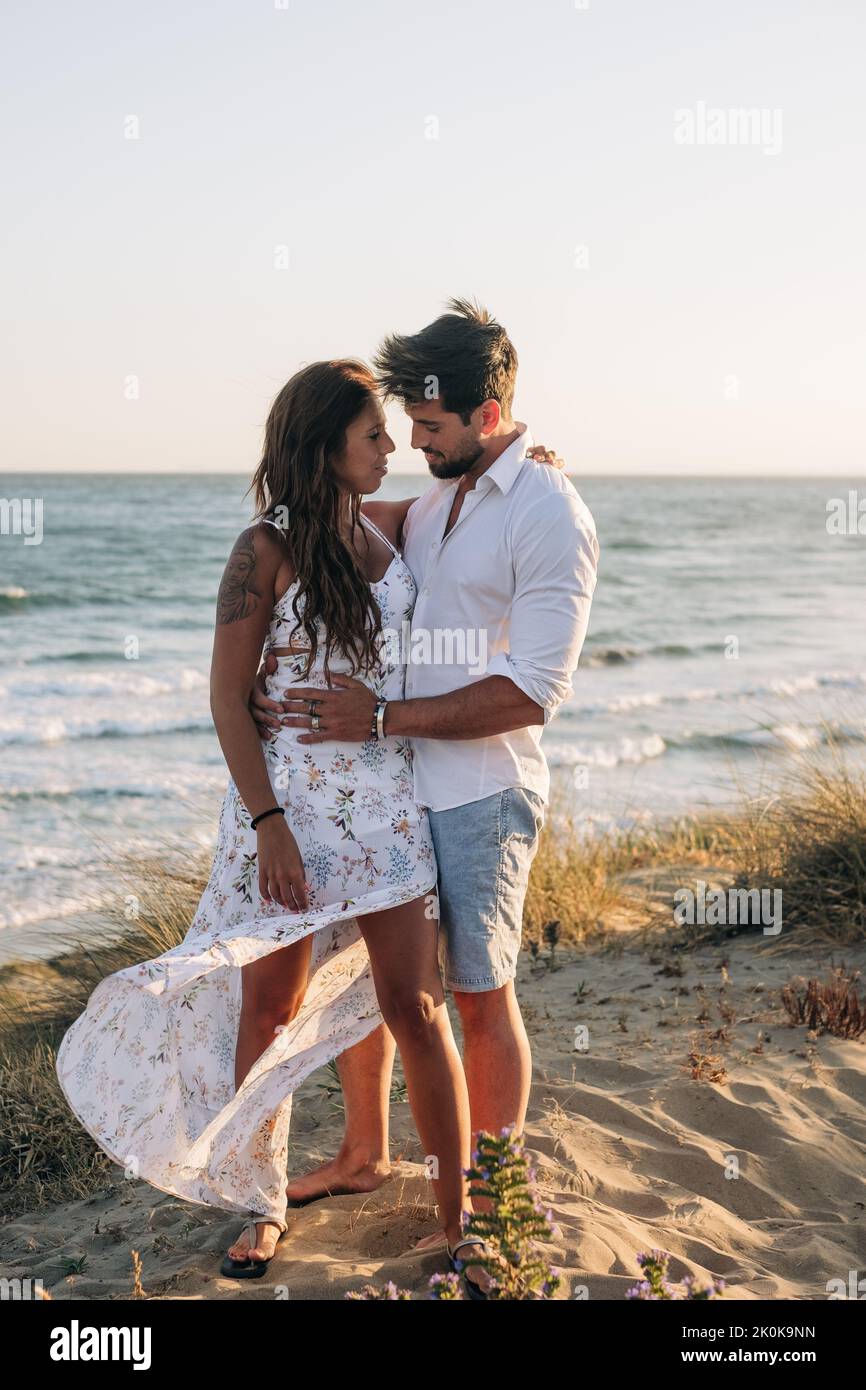 Happy young sunlit couple embracing on sandy beach by wavy ocean on ...