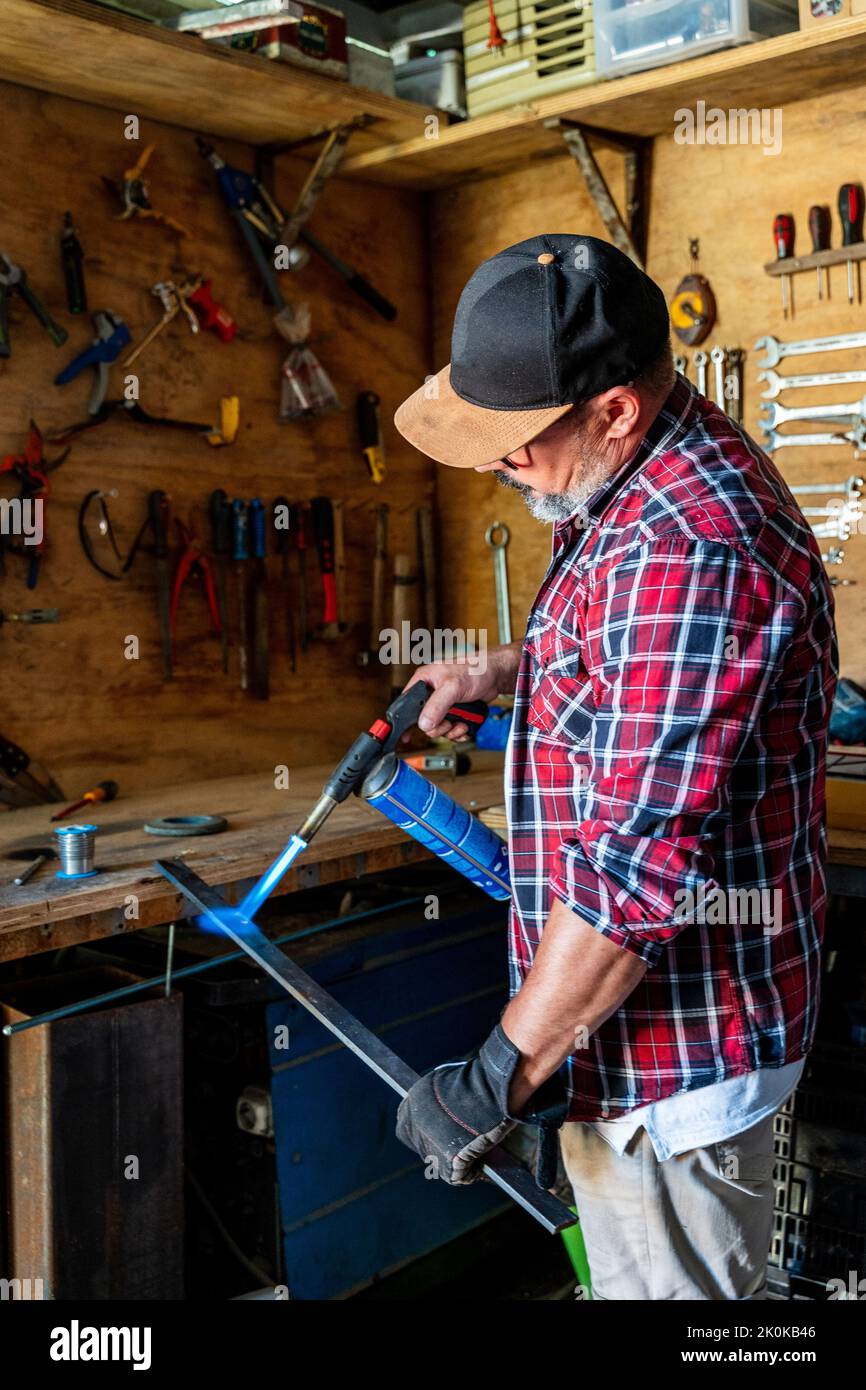 Middle aged casual carpenter in cap and gloves heating metal part with ...