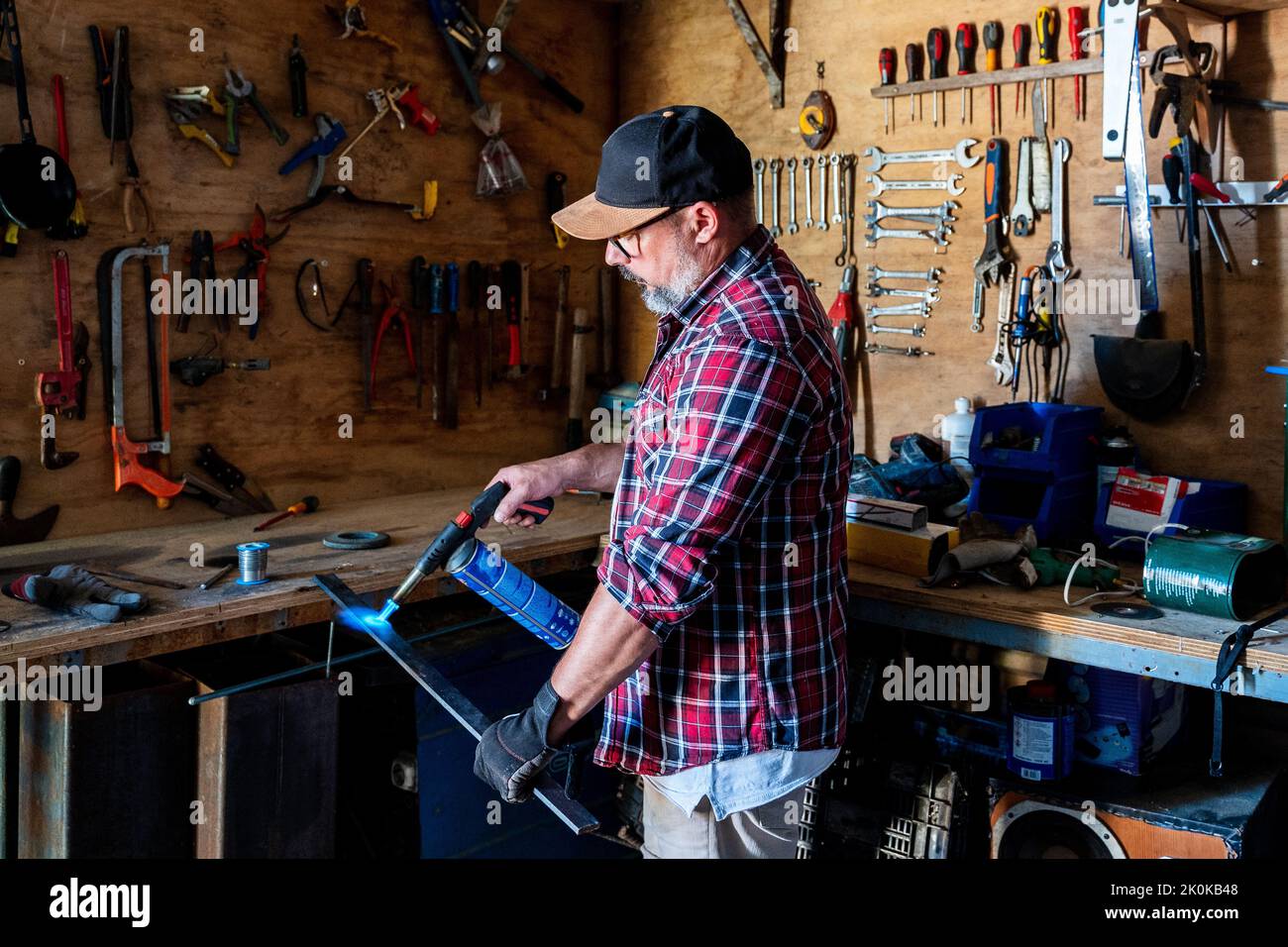 Middle aged casual carpenter in cap and gloves heating metal part with ...