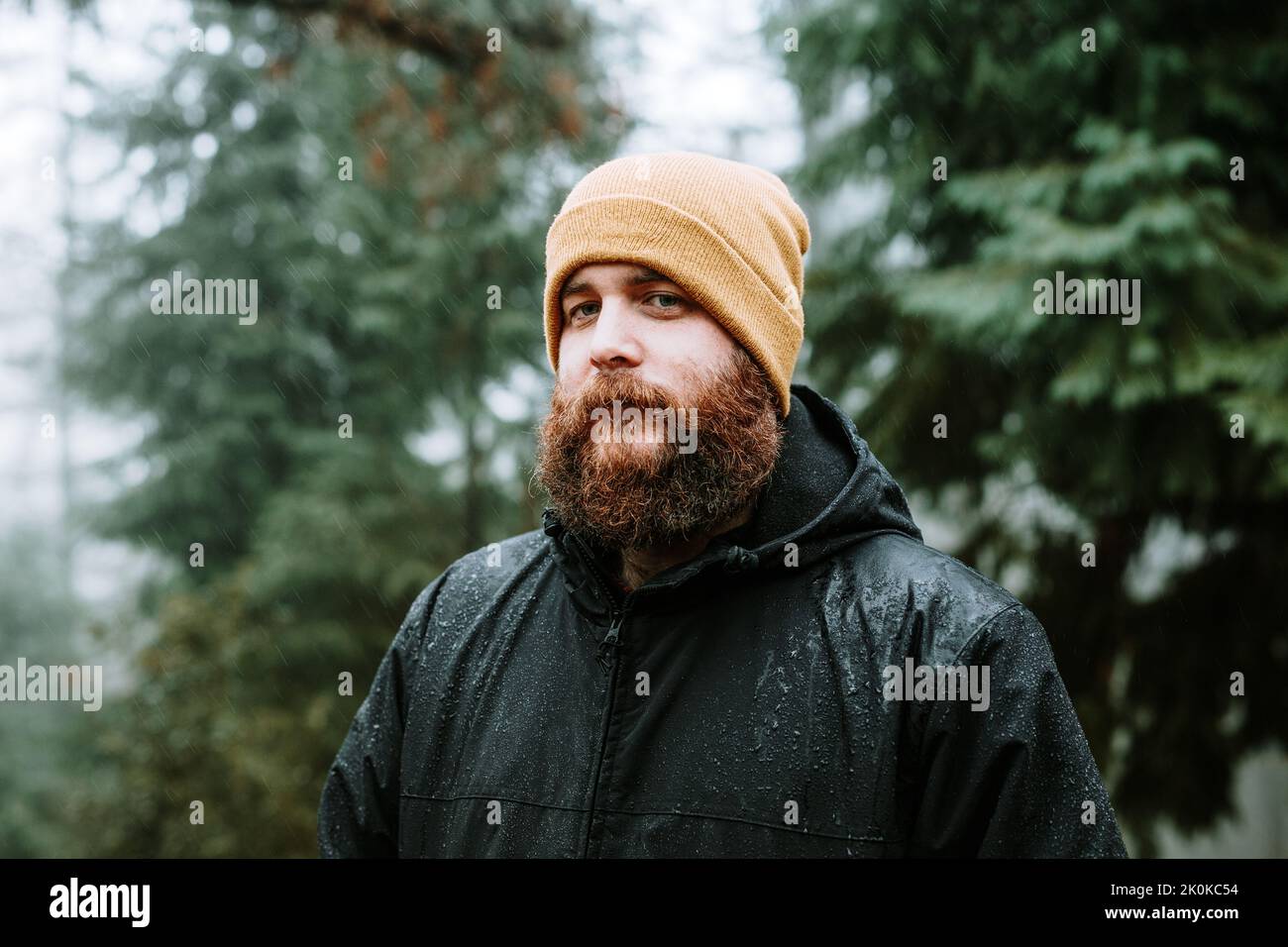 Thoughtful young man in coat with water drops and hat looking at camera ...