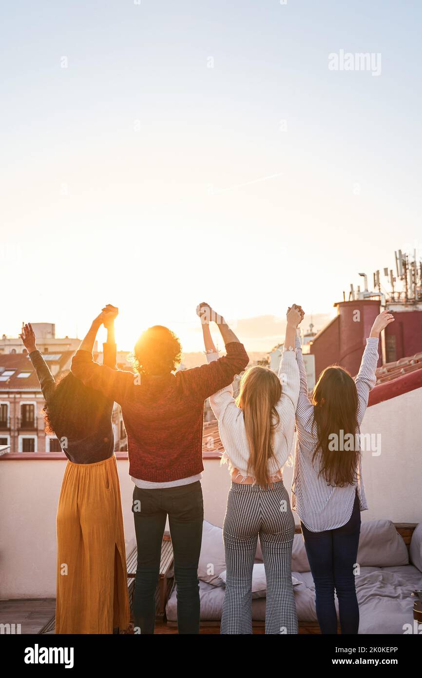 Back view faceless excited friends with arms raised in back lit wearing ...