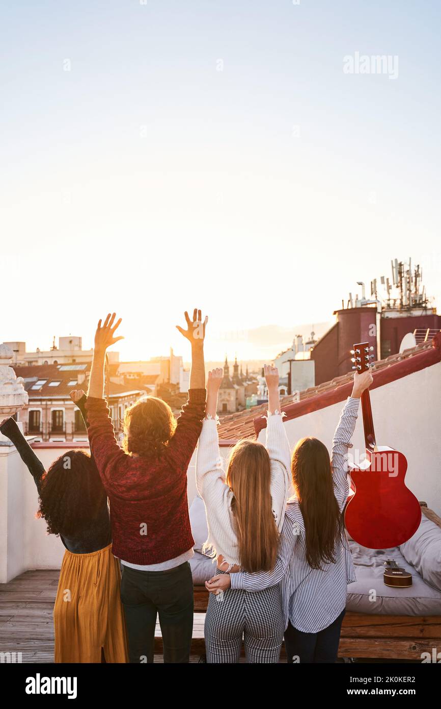 Back view faceless excited friends with arms raised in back lit wearing ...