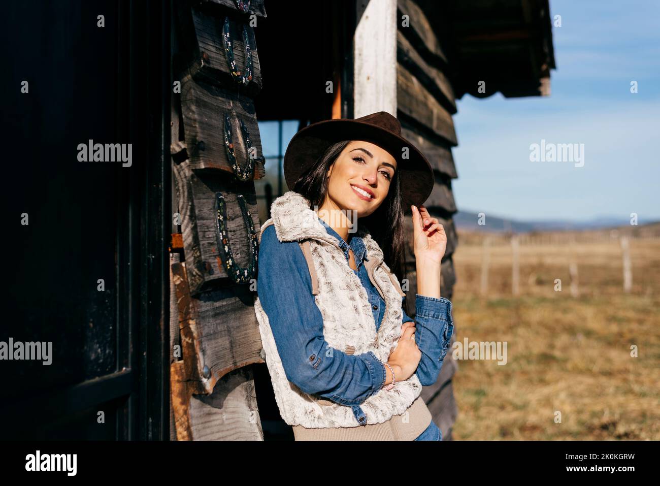 Beautiful brunette in cowboy and furry vest standing on porch of ranch ...