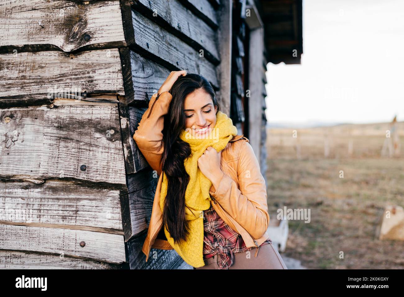 Stylish woman on ranch standing leaning on a wooden cabin looking down ...