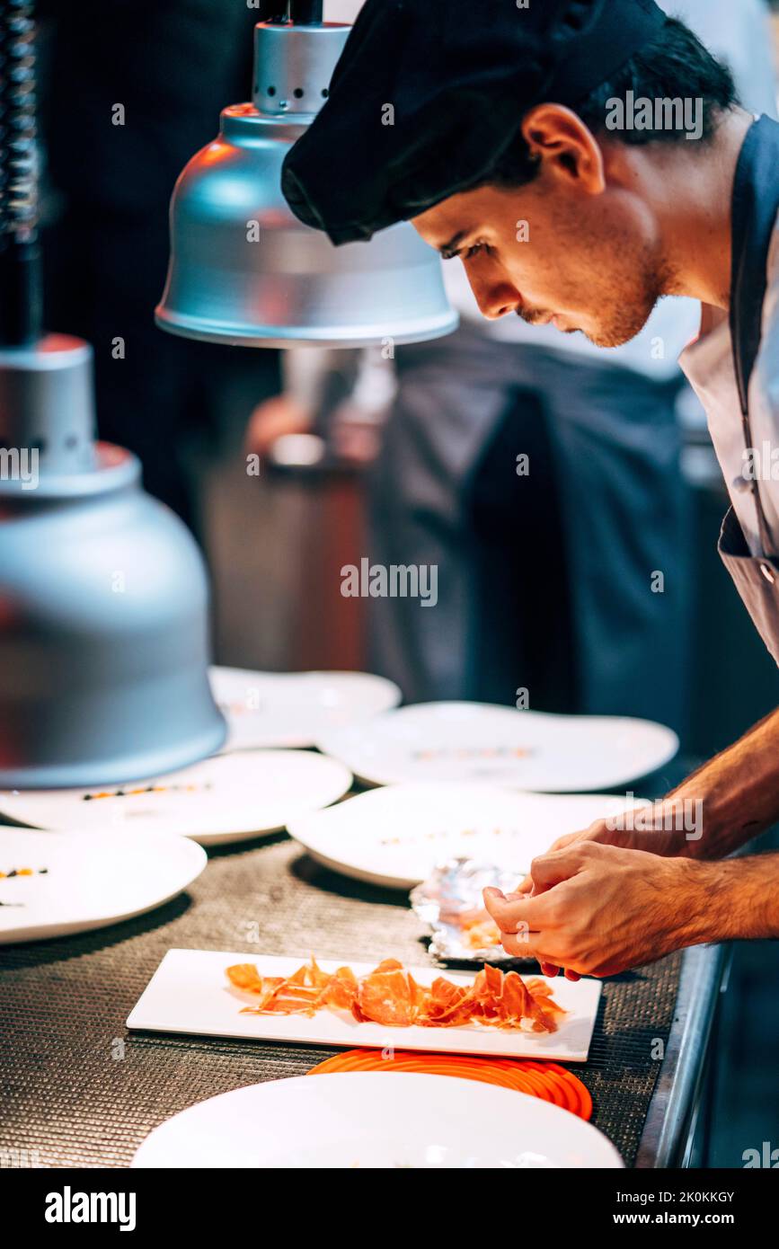 Side view of professional chef in hat serving ham in restaurant kitchen ...