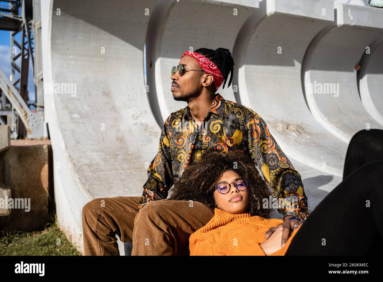 Black man sitting while woman with curly hair lies down in his lap looking away both wearing ...