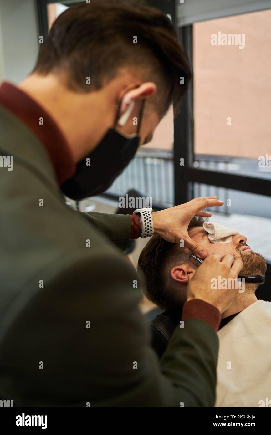 Back view of male barber in mask using razor and shaving beard of ...