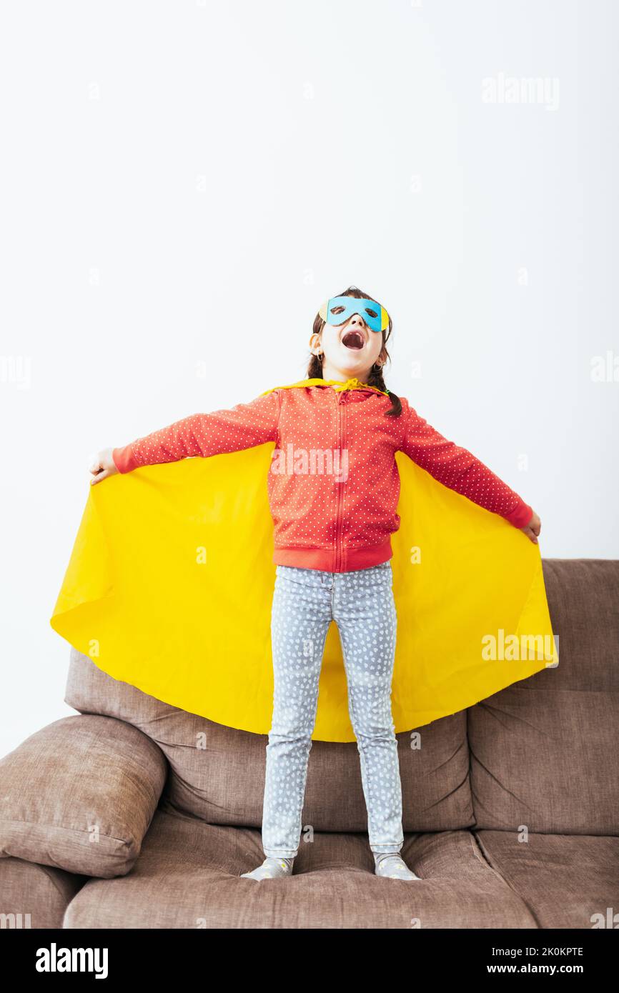Full body of confident kid in superhero cape and mask standing on sofa ...