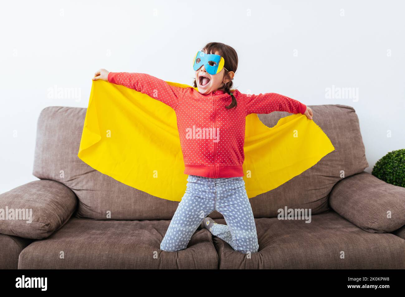 Confident kid in superhero cape and mask on sofa with the cloak open ...