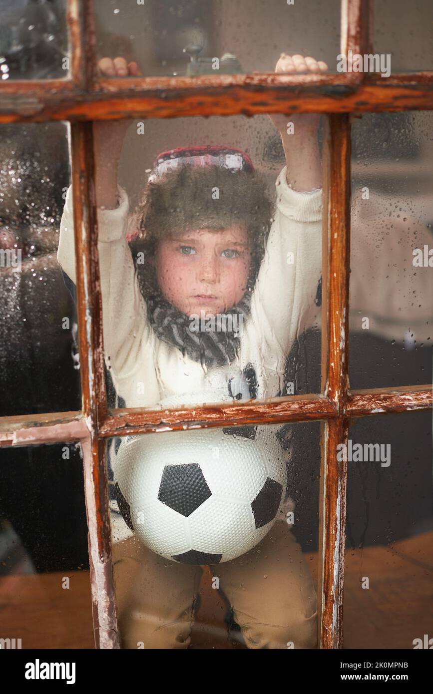 Will it ever stop raining. a sad little boy holding a soccer ball while ...
