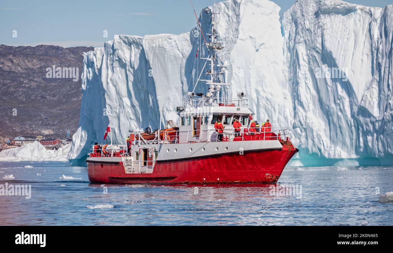 Bright red fishing ship carrying tourists in front of towering iceberg ...