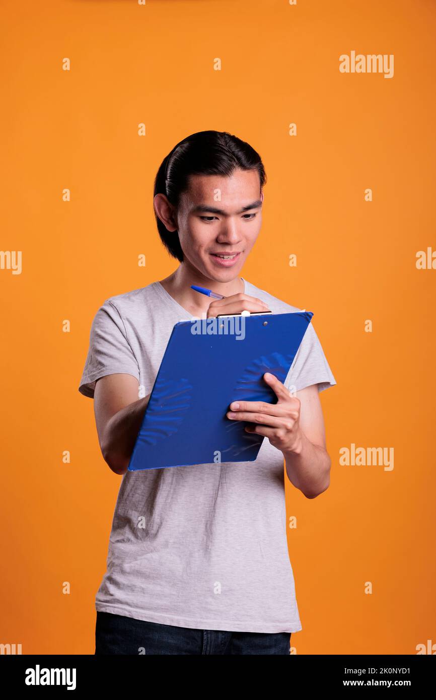 Smiling asian man taking notes in blue clipboard, student writing in ...