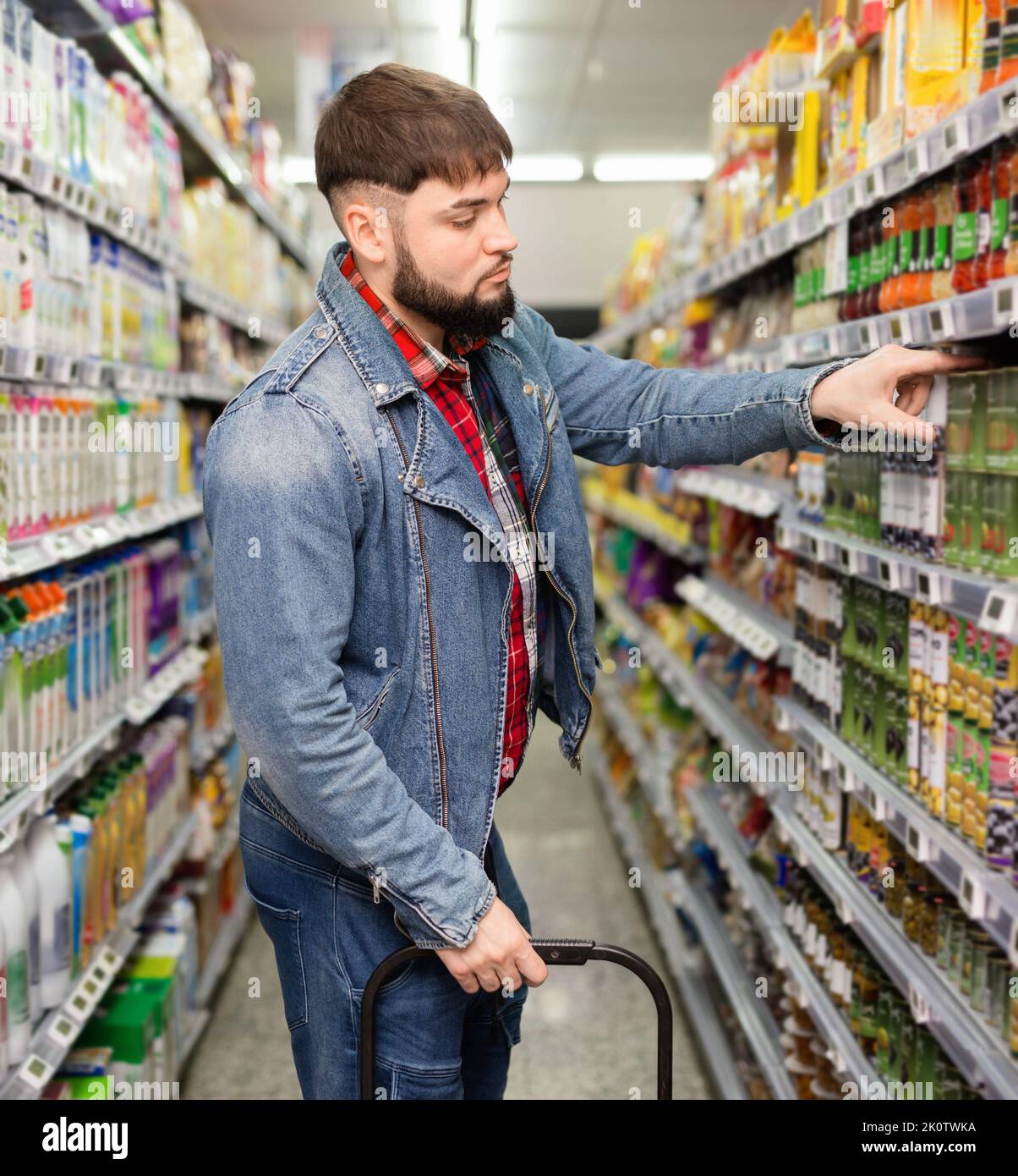Portrait of young man making purchases in grocery shop, choosing canned ...