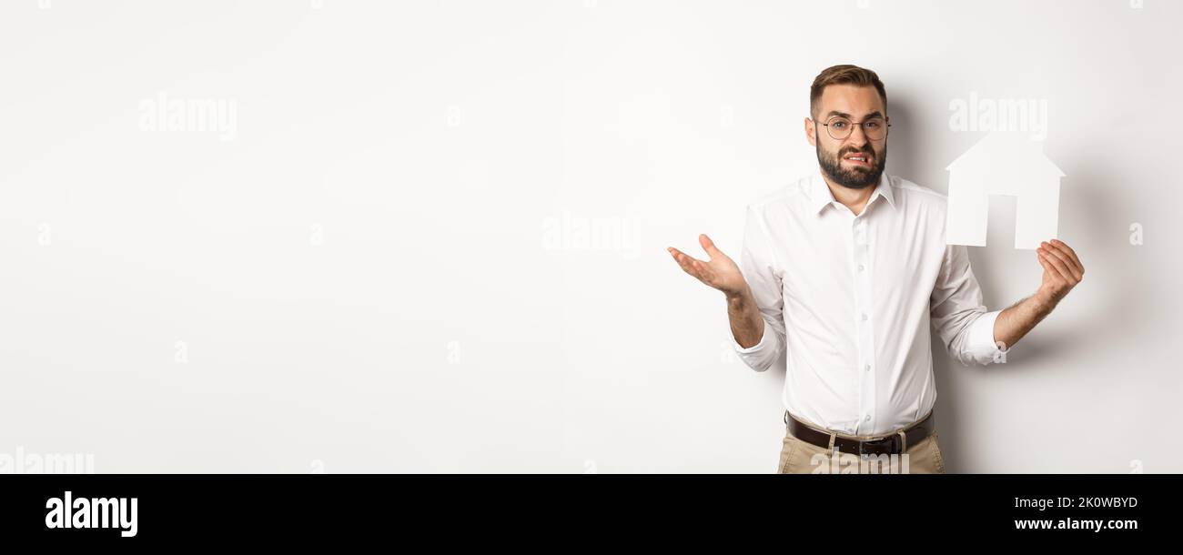 Real estate. Confused man shrugging, showing house paper model and ...