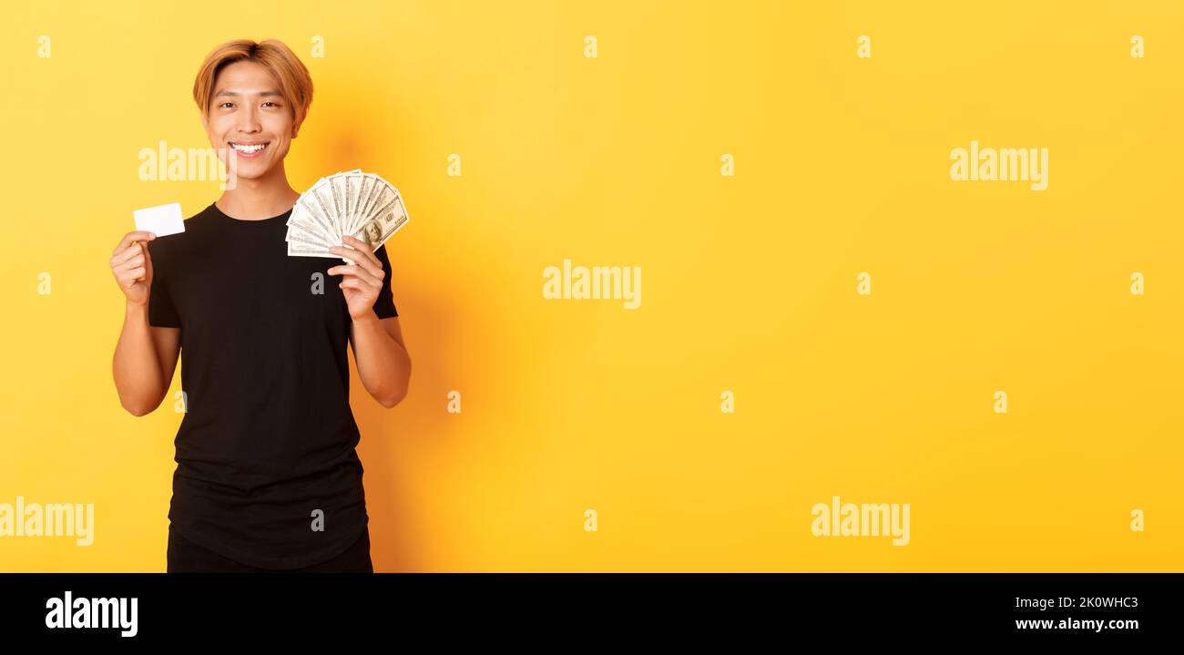 Pleased handsome asian guy showing money and credit card, smiling happy ...
