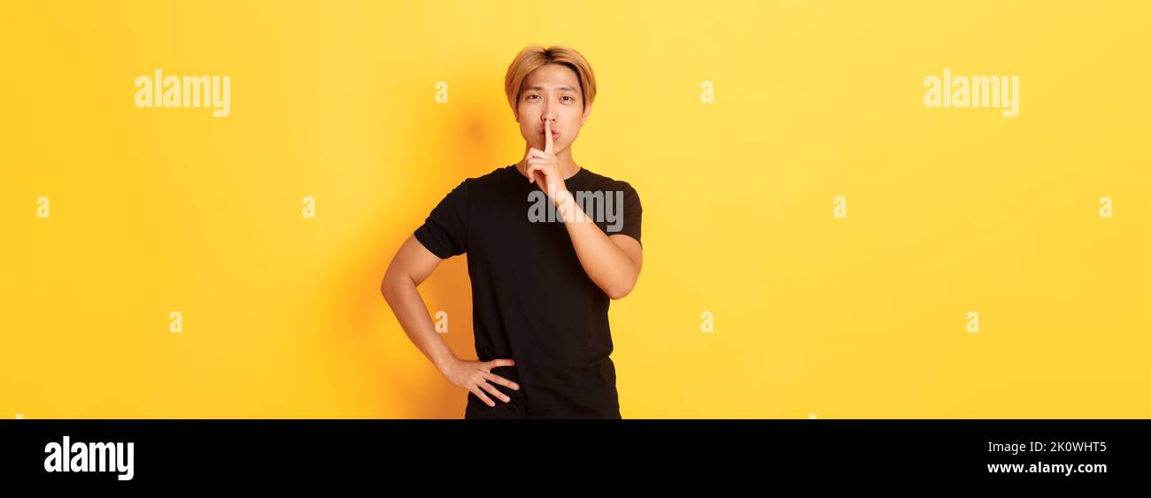 Portrait of attractive blond asian man shushing, press finger to lips ...