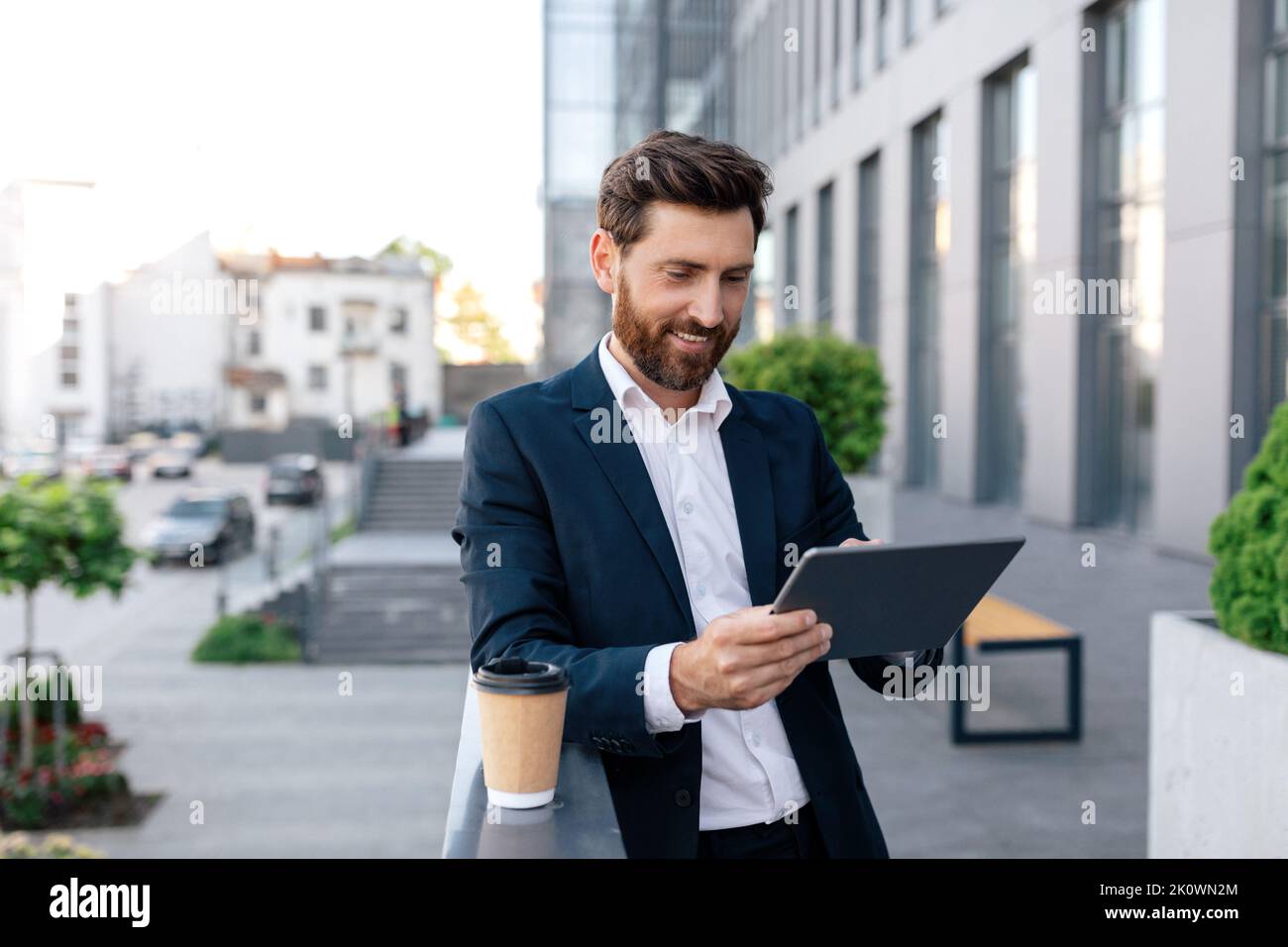 Happy confident young caucasian guy with beard in suit typing on tablet ...