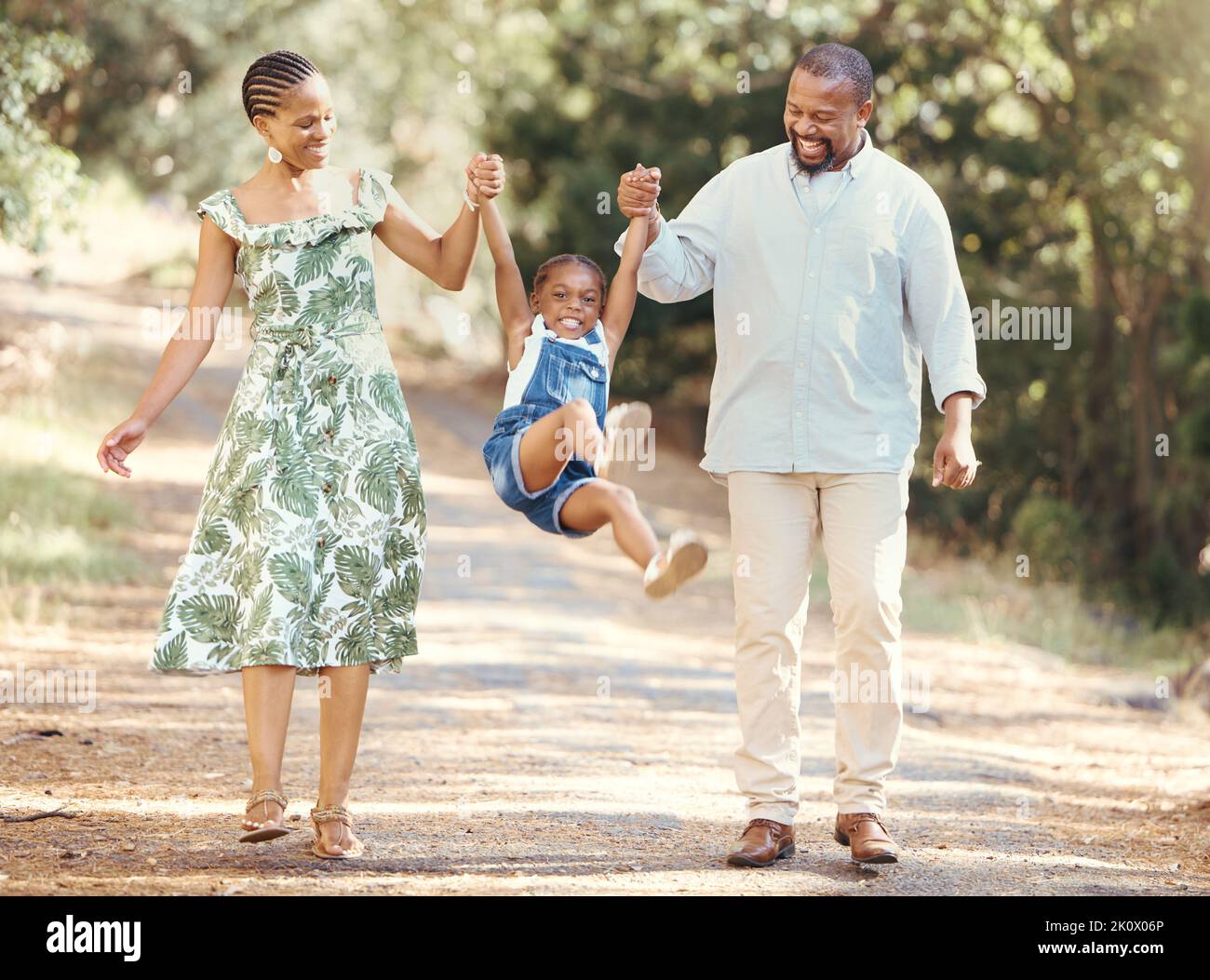 Happy parents swing girl at their hands in the forest during a walk in nature. Cheerful kid ...