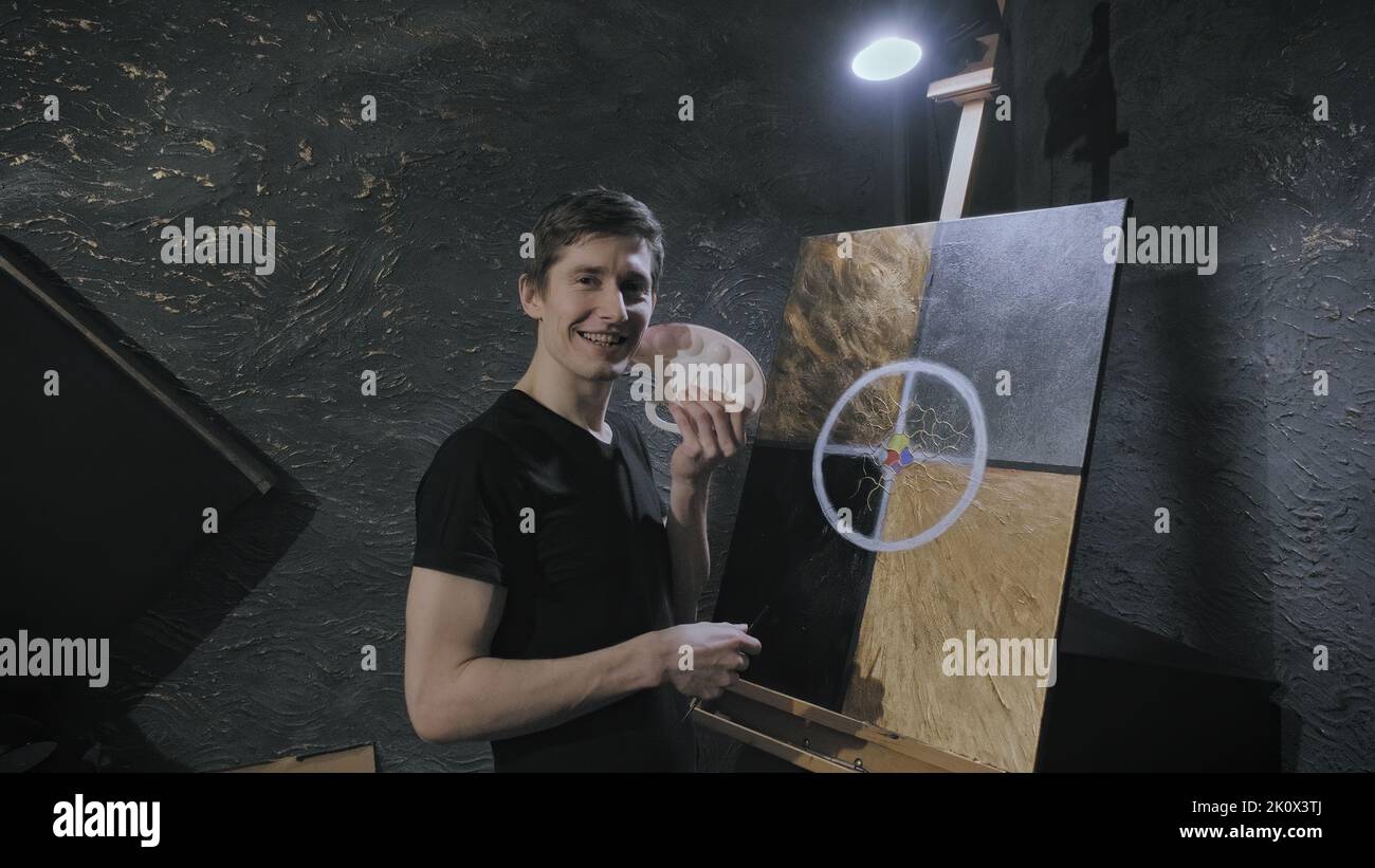 Smiling artist stands next to painting and holds palette with brush in ...