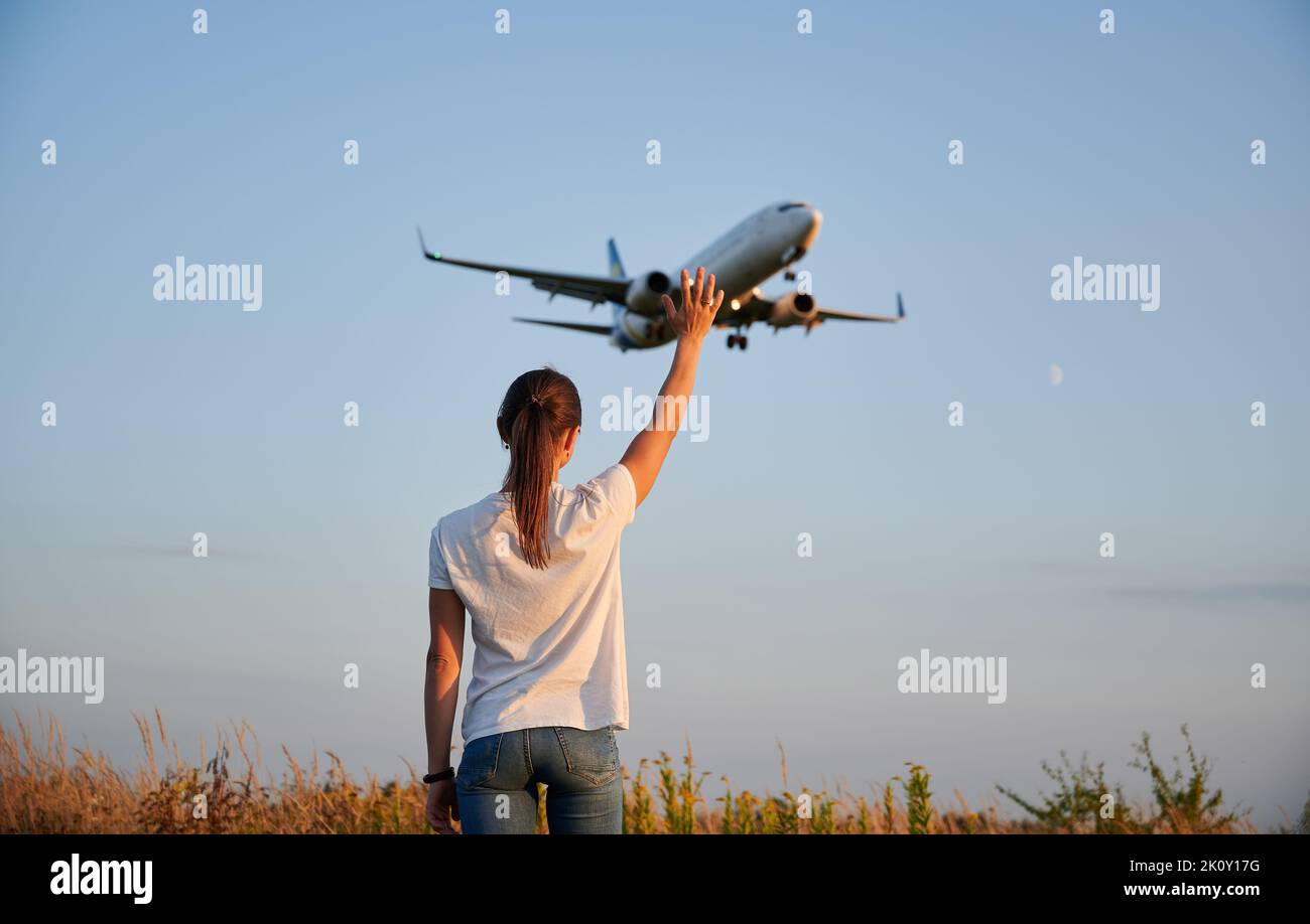 Back view of woman waving hand to flying commercial airplane in the sky ...