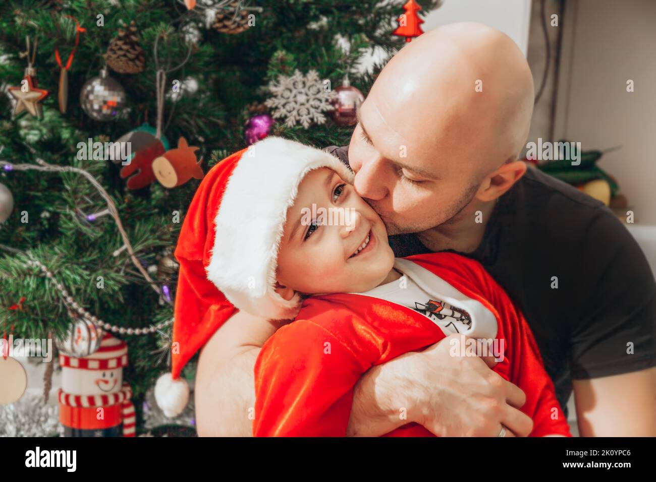 Dad kisses his son, dressed in a Santa costume, near the Christmas tree ...