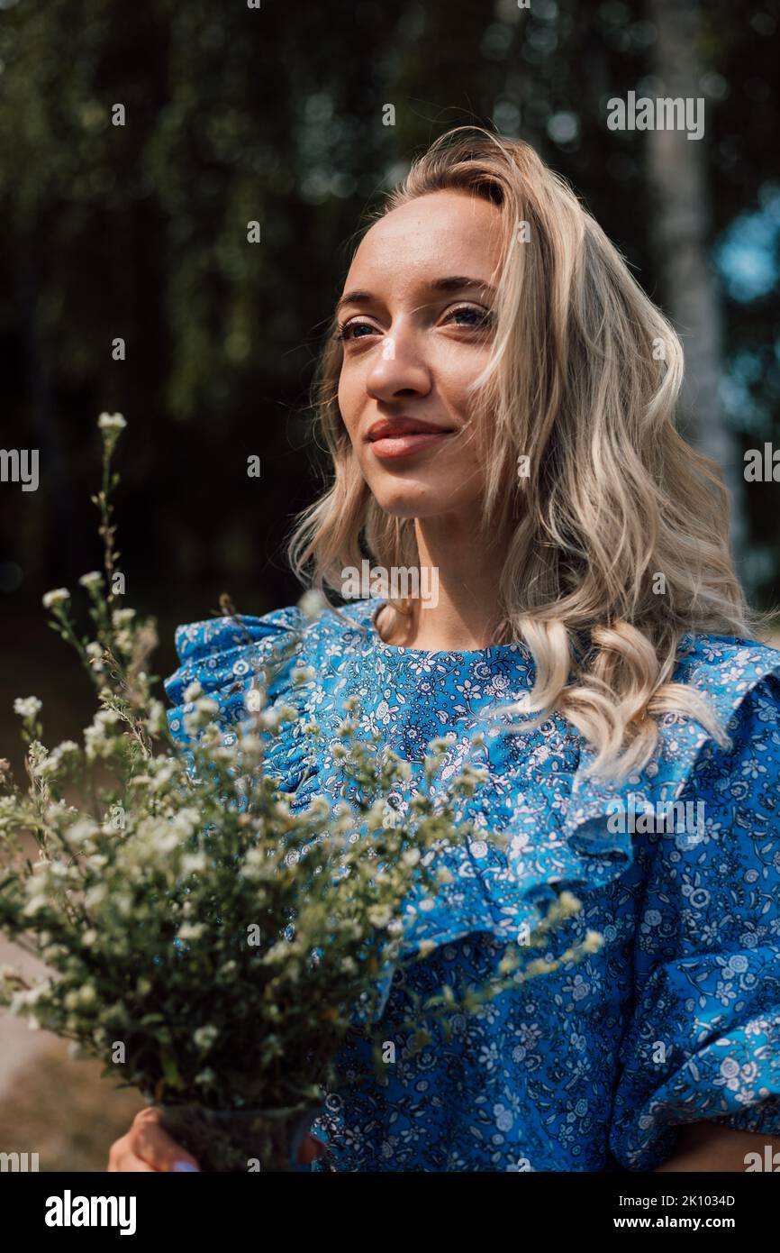 A young beautiful woman in a blue dress holds wild flowers in her hands ...