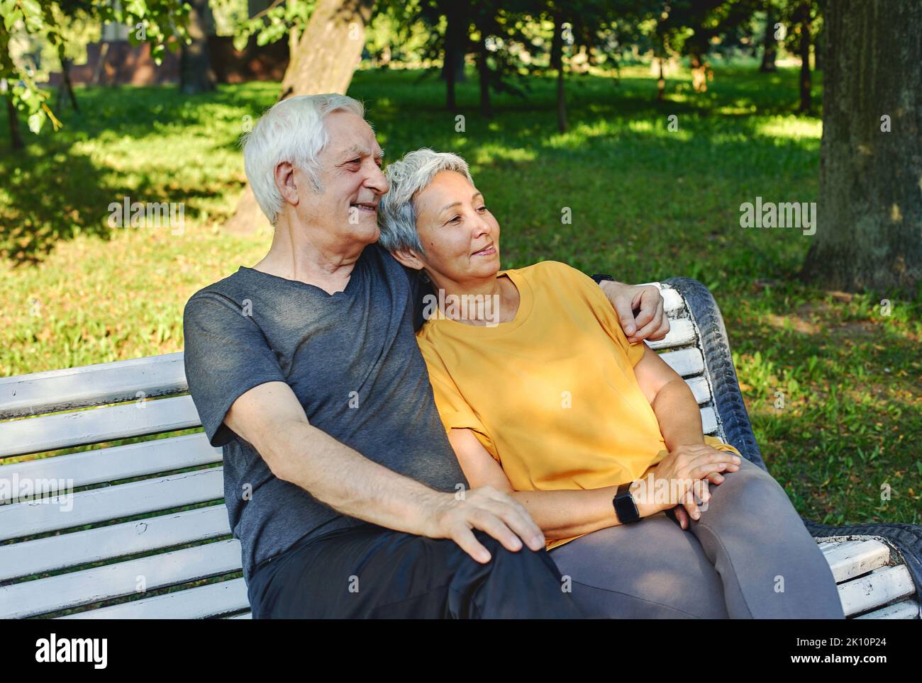 Pretty multi racial elderly grey hair couple resting sit on bench in park after stroll, morning ...