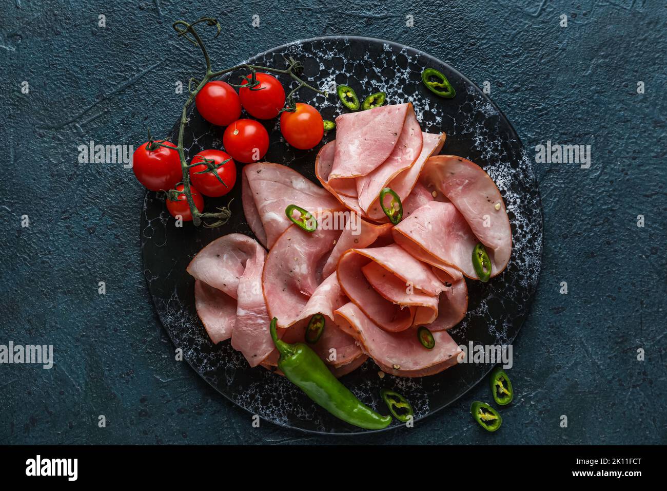 Board with slices of delicious ham and vegetables on dark color ...