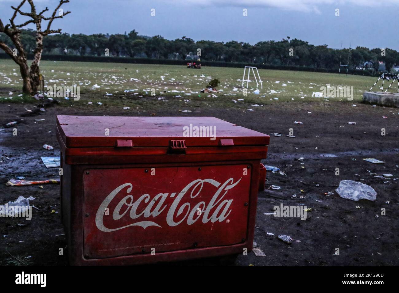 A Coca-Cola branded cooler box stands at a field littered with plastic ...